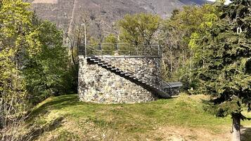 Uma escadaria de pedra leva a um corrimão de metal, cercada por árvores e uma paisagem montanhosa ao fundo.