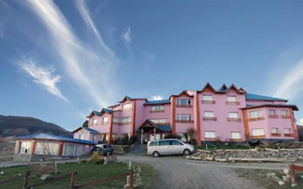 Um edifício rosa de vários andares com telhado azul, um carro estacionado e um prédio menor com telhado azul ao primeiro plano.