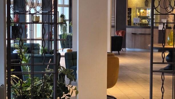 Um espaço interior com design moderno, apresentando uma área de estar com cadeiras e uma mesa, um balcão de recepção e vista através de um painel de vidro com plantas.