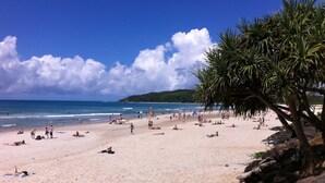 Uma praia com pessoas tomando sol e nadando, uma torre de salva-vidas e um litoral rochoso com palmeiras.
