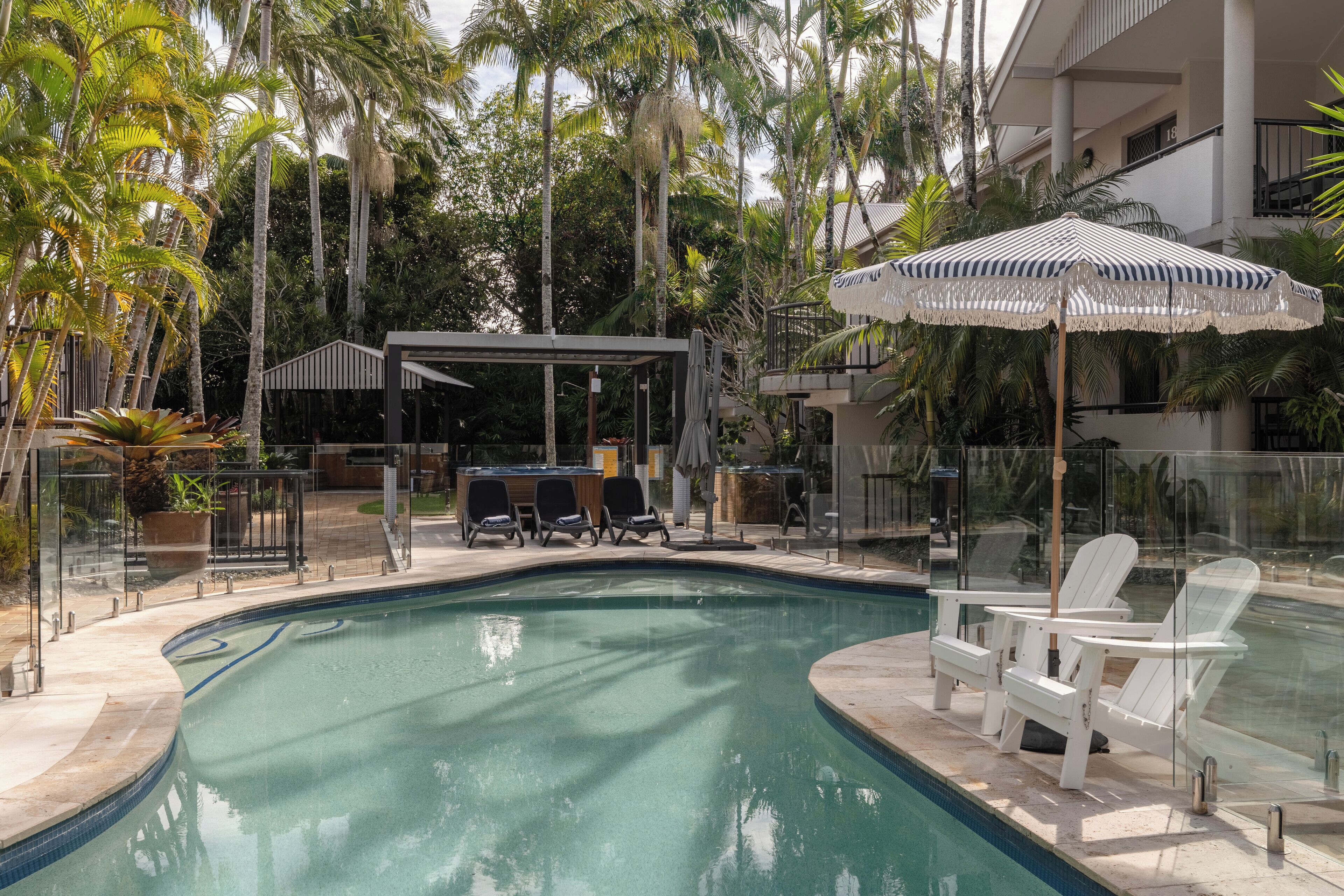 Piscina com espreguiçadeiras e área de estar coberta, cercada por palmeiras e um edifício com sacadas.