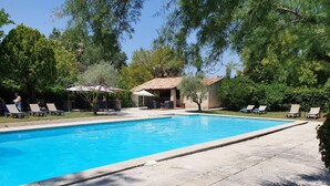 Piscina retangular com espreguiçadeiras e guarda-sóis, rodeada por um deck ladrilhado, com árvores e um edifício ao fundo.