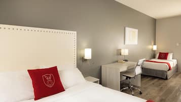 Quarto de hotel com uma cama grande, uma escrivaninha e uma cama menor. Há uma luminária de mesa, uma cadeira e um travesseiro vermelho com um brasão.