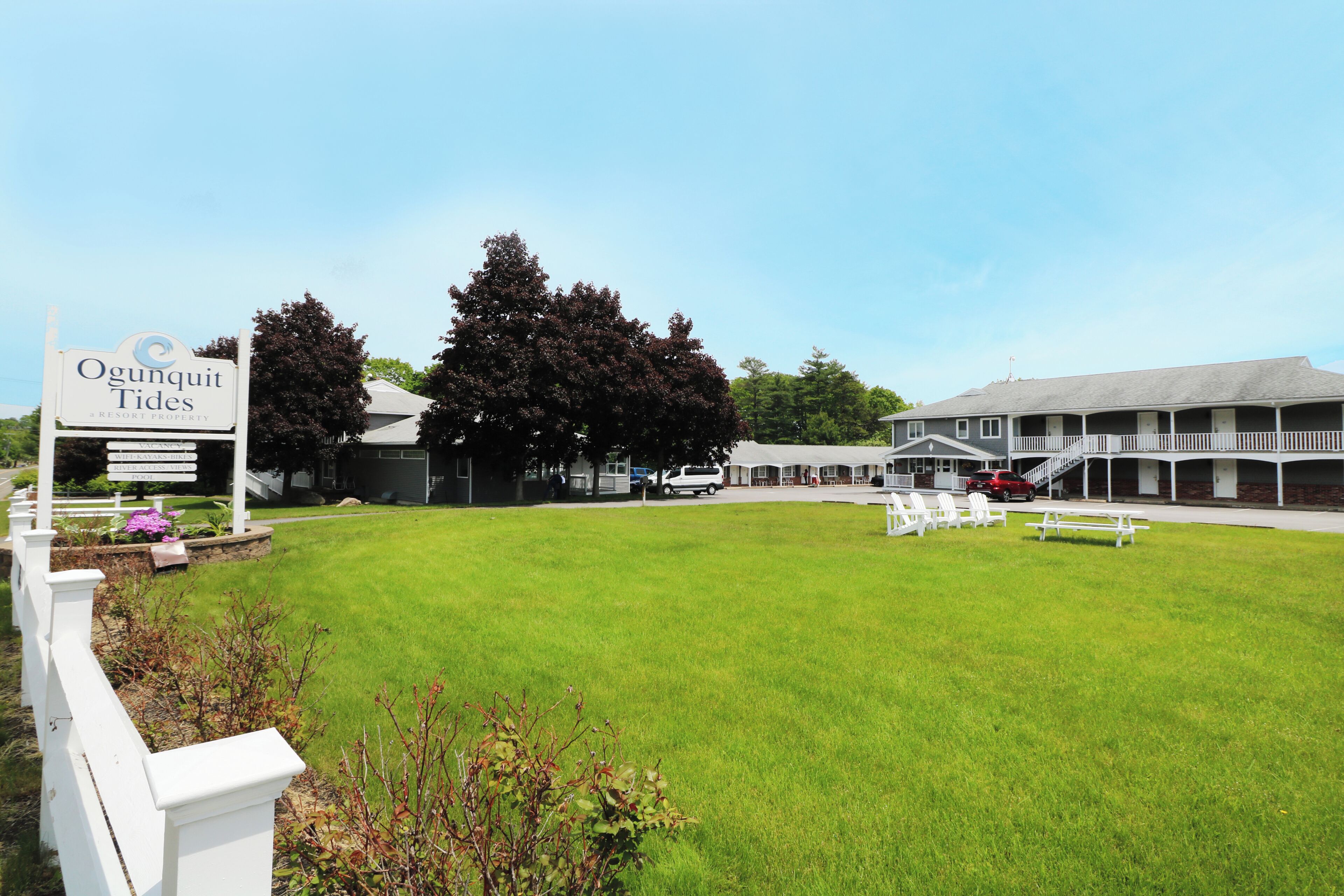 Uma área gramada com uma placa para Ogunquit Tides Resort Property, um edifício com varanda coberta e um cercado branco.