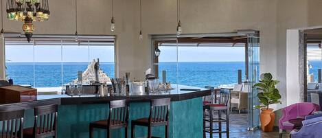 Um bar com vista para o oceano, banquetas e uma grande planta em vaso.