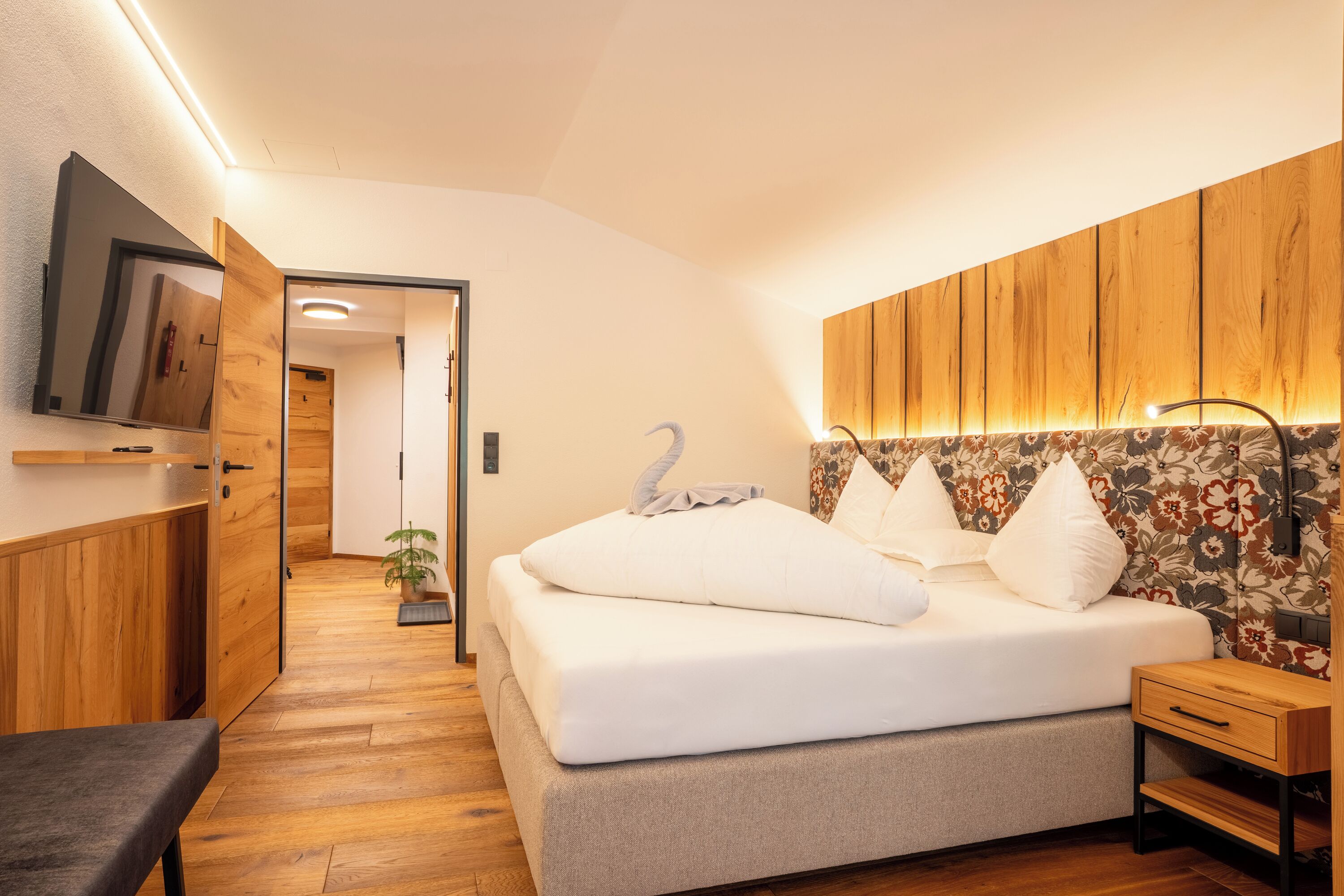 Quarto com cama, mesa de cabeceira, guarda-roupa de madeira e televisão na parede.