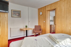 Quarto de hotel com duas camas, uma cadeira de madeira, uma mesa pequena com uma planta, um quadro na parede e um guarda-roupa de madeira.