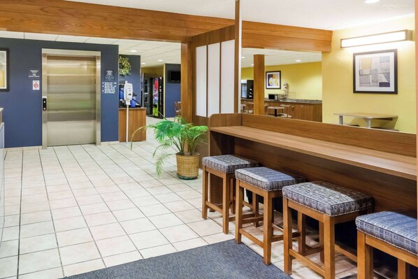 Um saguão de hotel com balcão de recepção, banquetas de madeira, um vaso de planta e um elevador.