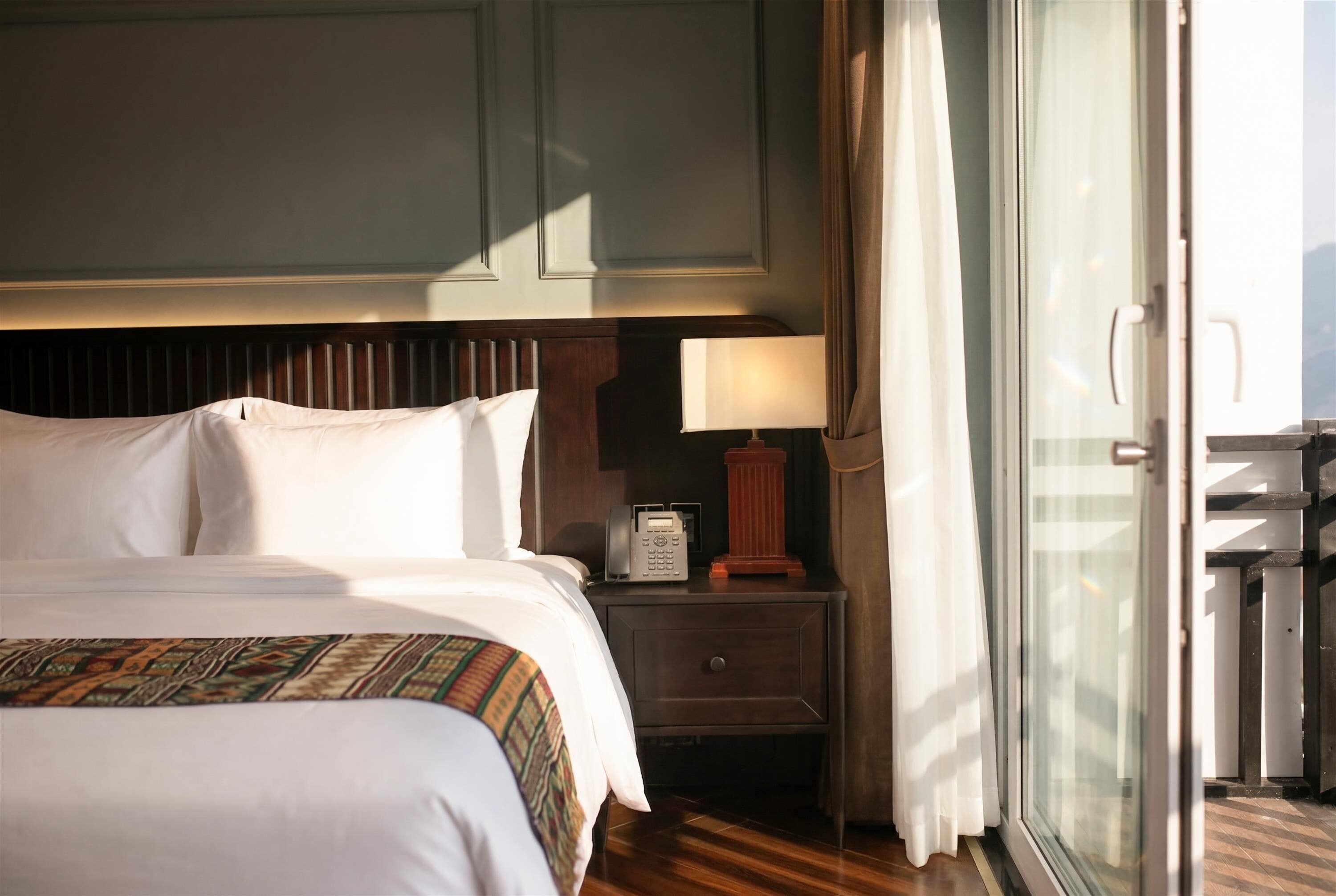 Quarto de hotel com uma cama, uma mesa de cabeceira com abajur, telefone e vista através de uma porta de vidro.