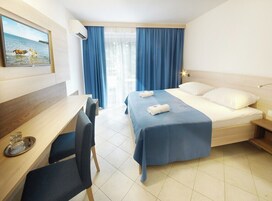 Quarto de hotel com cama, escrivaninha e cadeira. Há uma TV fixada na parede, e uma janela com cortinas.
