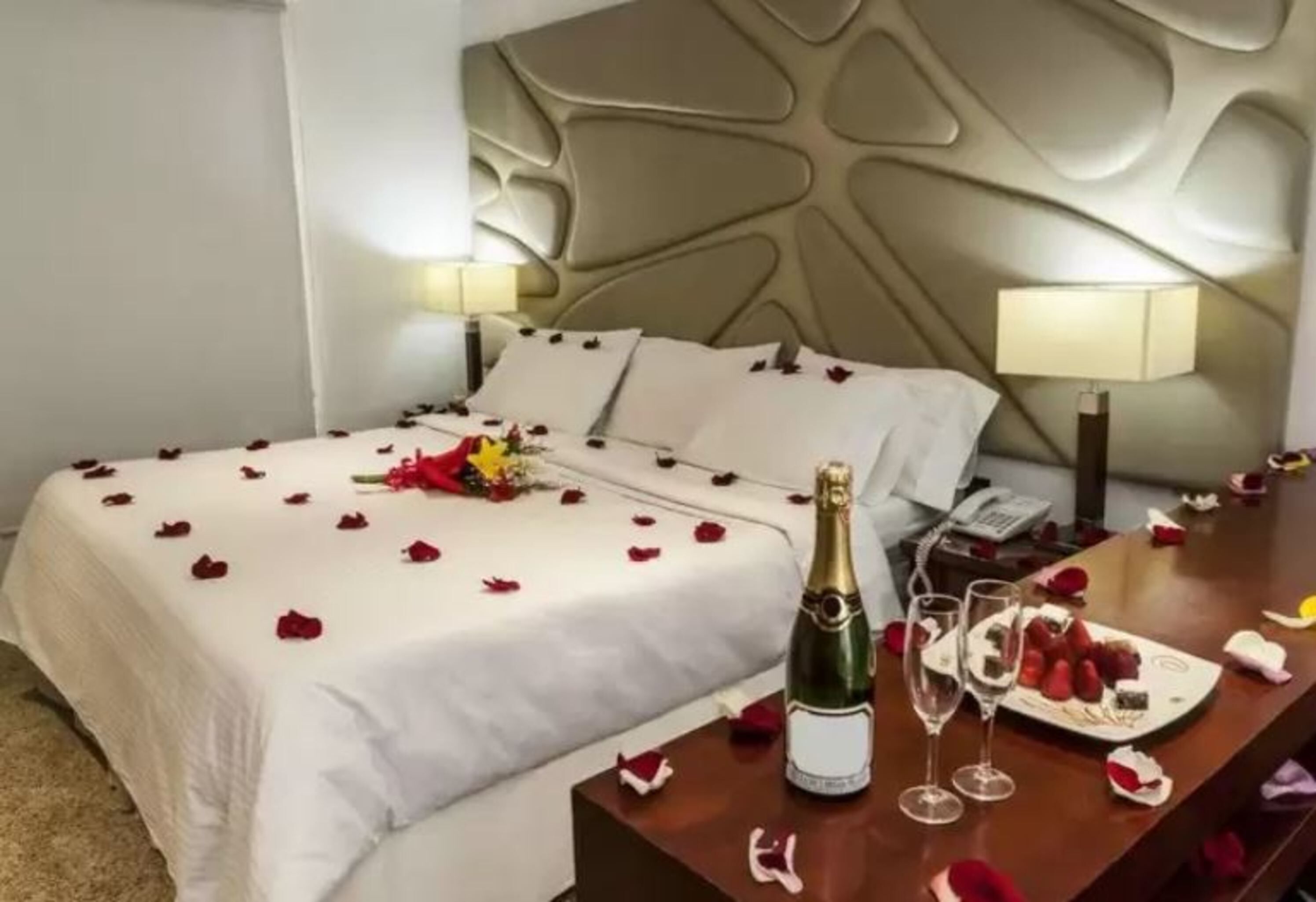 Quarto de hotel com cama decorada com pétalas de rosas, uma garrafa de champanhe e uma tábua de frutas.
