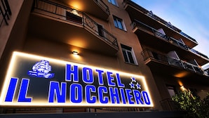 Placa de hotel com letras azuis iluminadas que dizem 'HOTEL IL NOCCIERO' ao fundo de uma fachada de prédio.