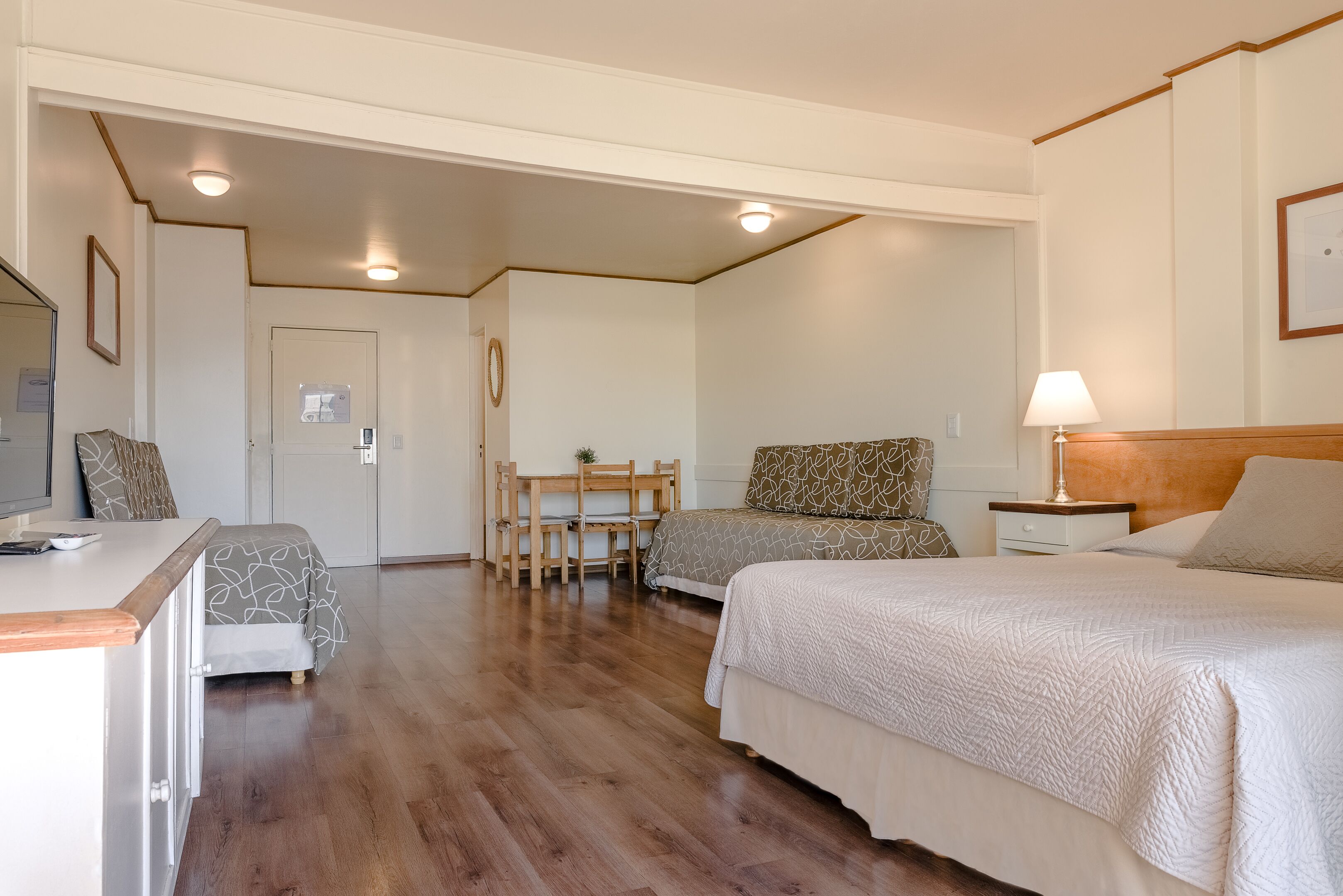 Quarto de hotel com cama, mesa de cabeceira, luminária, escrivaninha, cadeira e uma pequena mesa com uma planta.