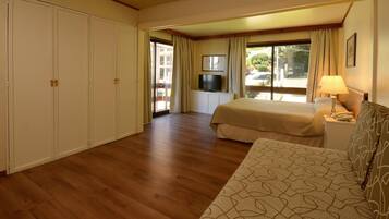 Quarto de hotel com chão de madeira, cama com colcha bege, sofá com almofada estampada, televisão sobre um móvel e janela com cortinas.