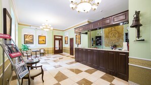 Um hall de hotel com piso xadrez, balcão de recepção em madeira e um candelabro.