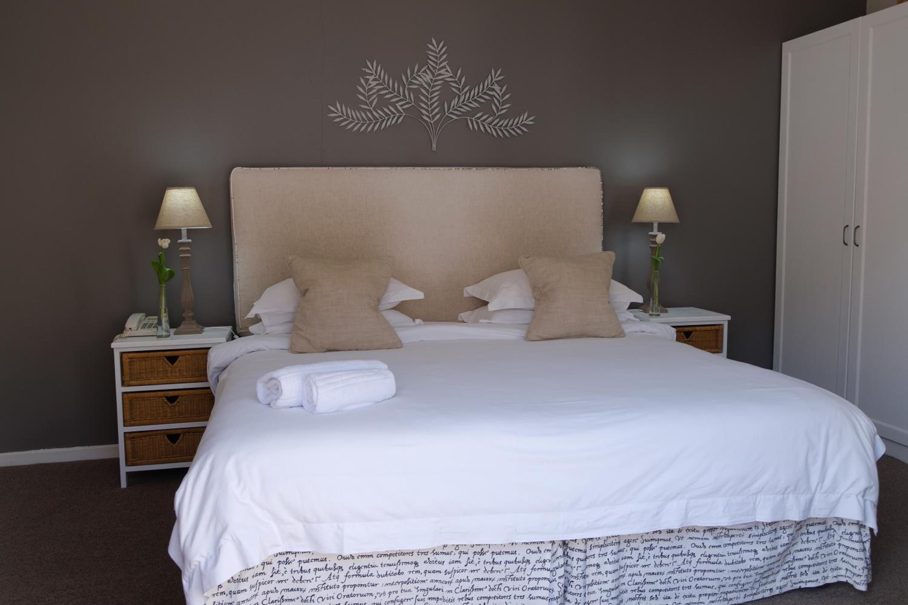 Uma cama bem arrumada com roupa de cama branca e duas almofadas bege, uma cabeceira branca e duas mesas de cabeceira com abajures e um vaso de flores.