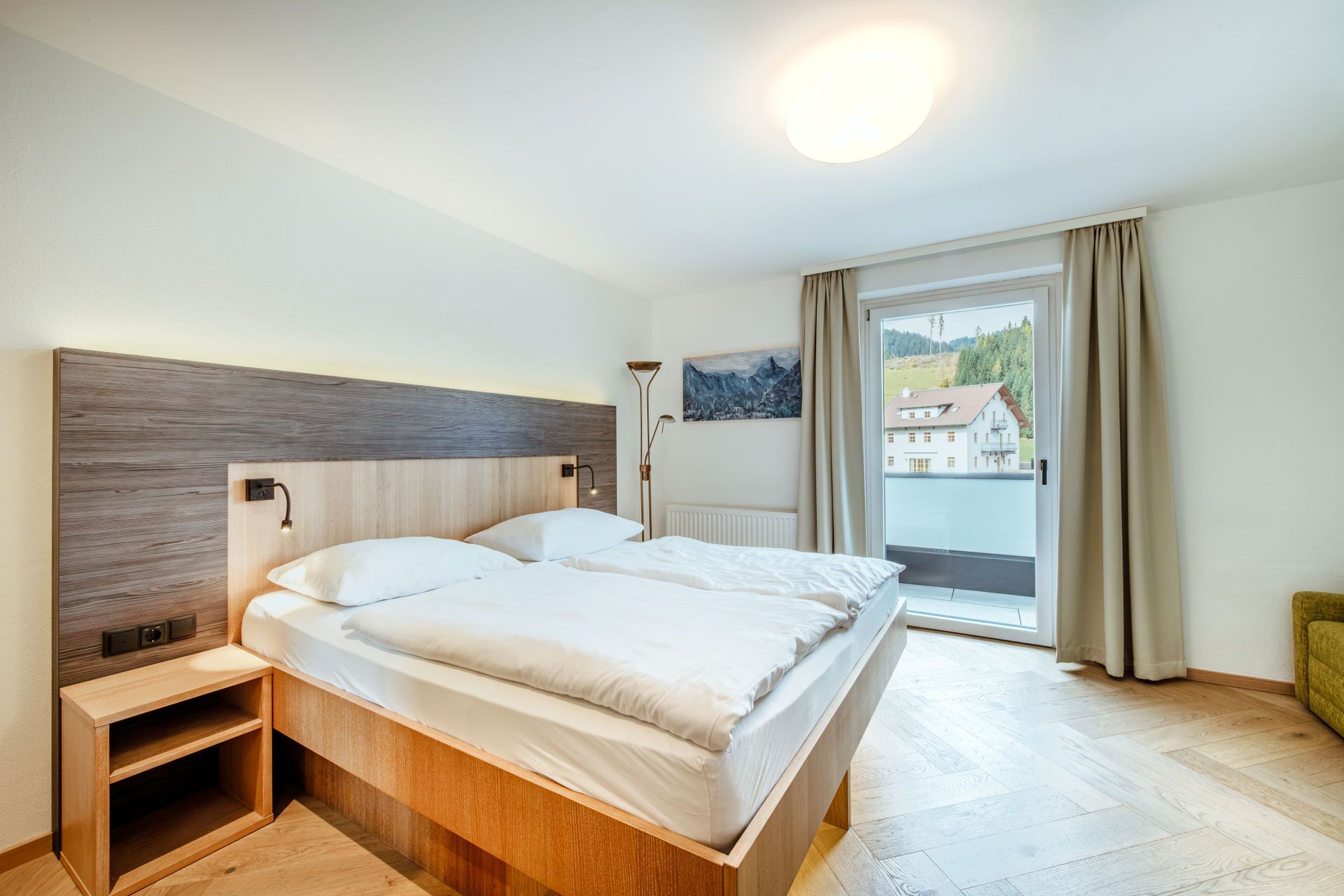 Quarto de hotel moderno com cabeceira de madeira, uma cama grande, mesa de cabeceira, abajur e vista para uma paisagem montanhosa pela janela.