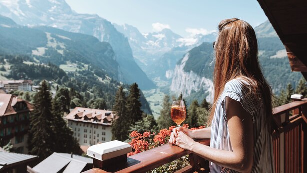 Uma pessoa segurando um copo com bebida, apreciando a vista de uma paisagem montanhosa com edifícios no vale.