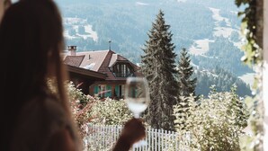 Uma pessoa segurando uma taça de vinho, olhando para uma paisagem montanhosa através de uma janela com vista para uma casa e uma cerca.
