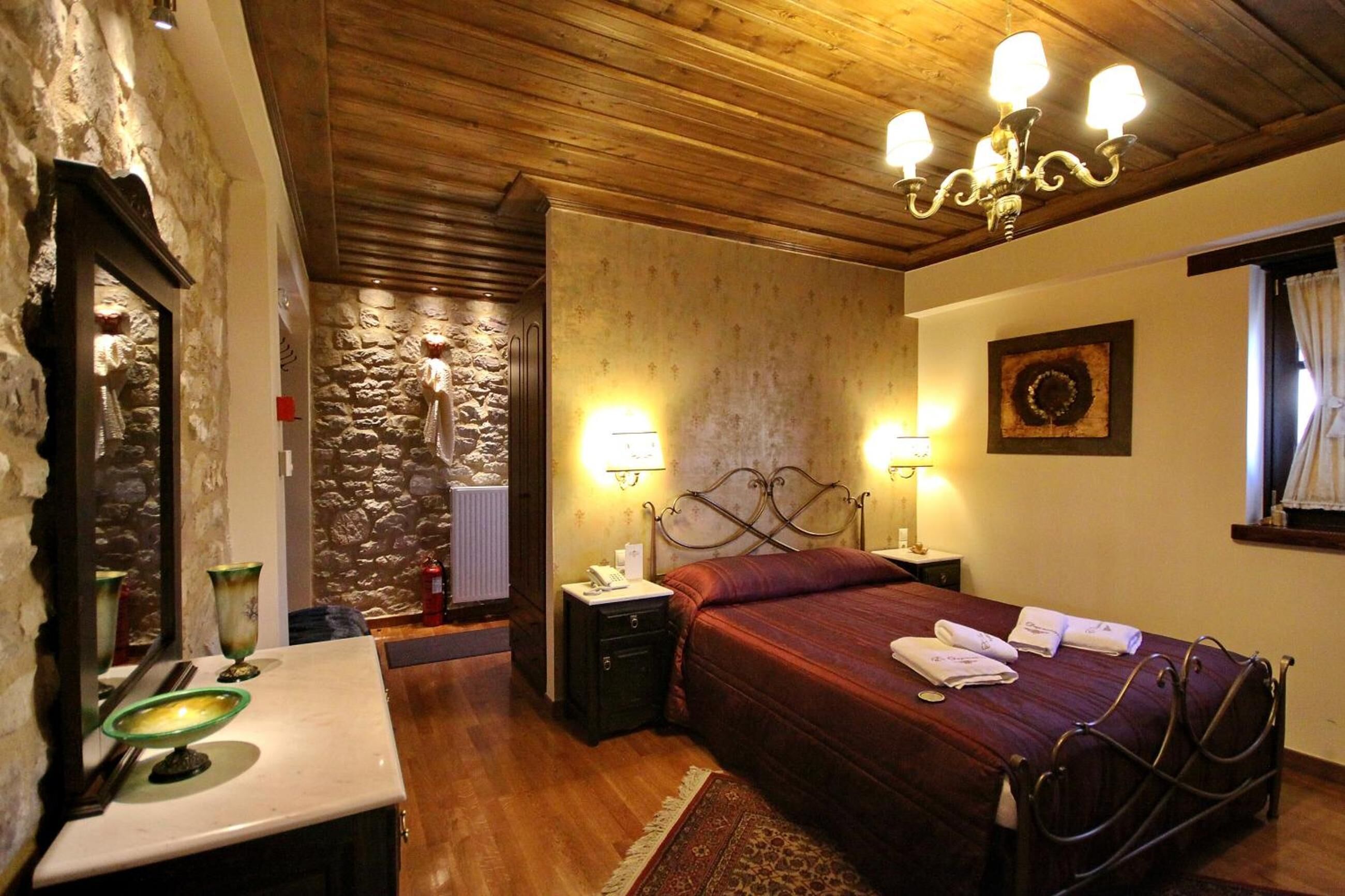 Quarto com teto de madeira, parede de pedra, duas luminárias de cabeceira, cama com colcha vermelha, mesa de cabeceira, espelho e janela com cortinas.