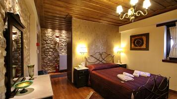 Quarto com teto de madeira, parede de pedra, duas luminárias de cabeceira, cama com colcha vermelha, mesa de cabeceira, espelho e janela com cortinas.