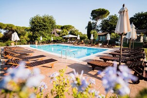 Área de piscina com espreguiçadeiras e guarda-sóis, cercada por vegetação e um edifício ao fundo.