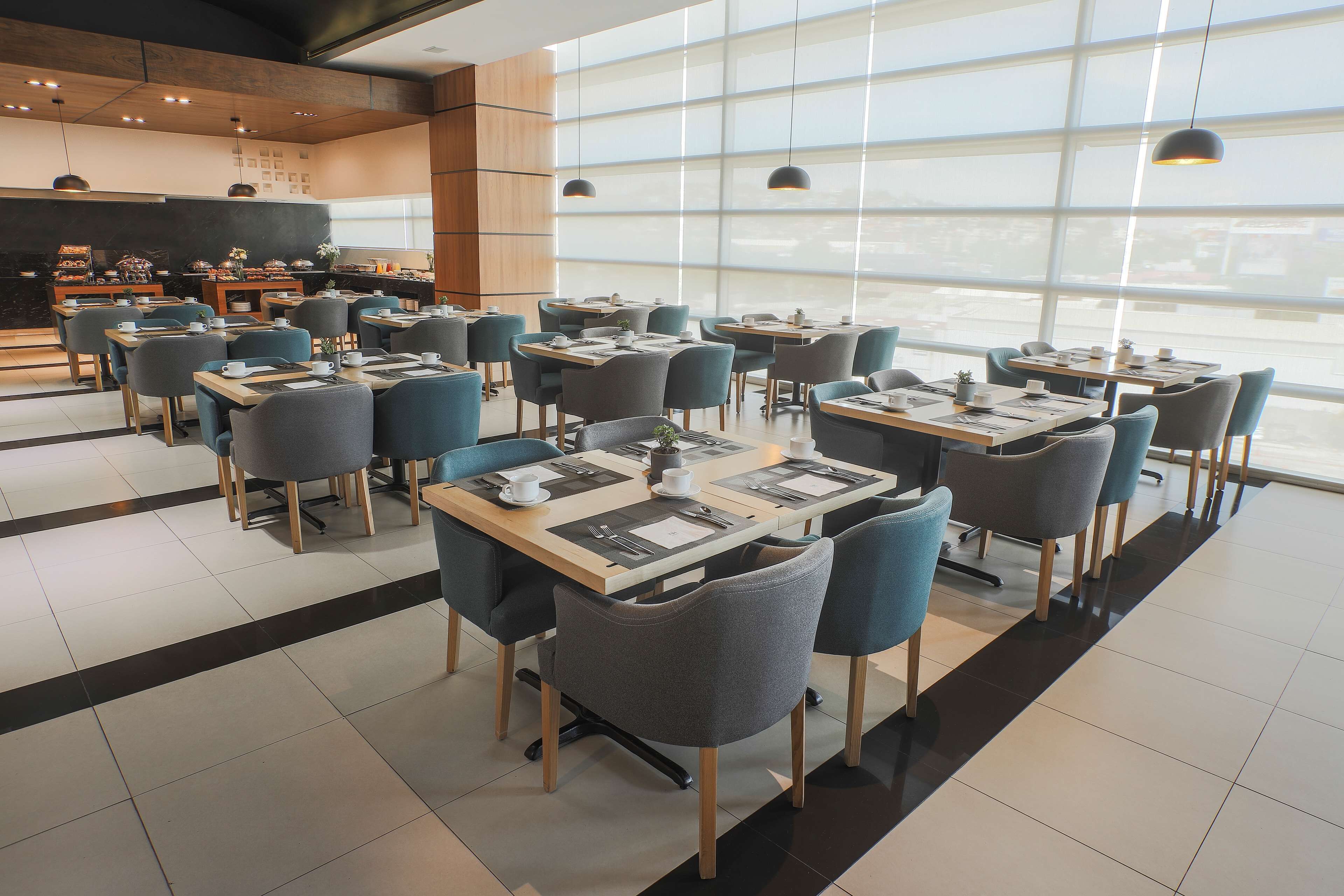 Um restaurante moderno com várias mesas e cadeiras, grandes janelas e uma área de cozinha ao fundo.