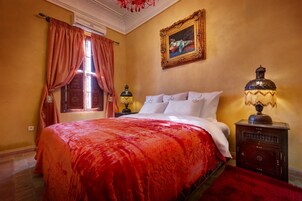 Quarto de hotel com uma cama grande, uma mesa de cabeceira com abajur, um quadro na parede e uma janela com cortinas.
