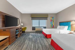 Quarto de hotel com uma televisão de tela plana grande, uma escrivaninha com gavetas, uma cadeira, duas camas com roupa de cama vermelha e branca, uma janela com vista e um ar-condicionado.