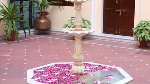 Uma fonte com pétalas de flores rosa em um pátio com piso de azulejos, rodeado por plantas em vasos e portas tradicionais.