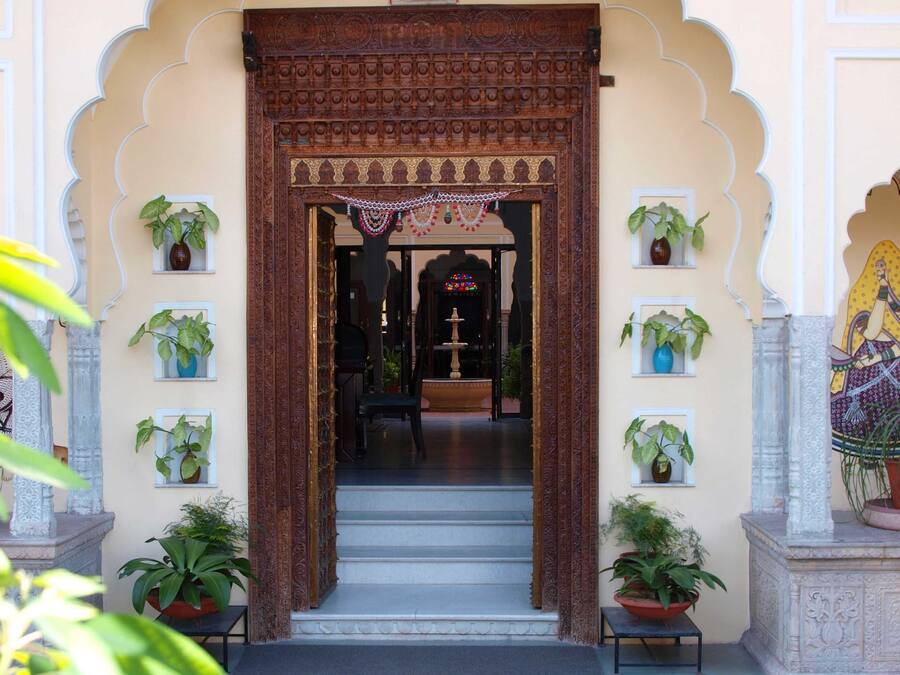 Uma entrada ornamentada com um arco decorativo, plantas em vasos e uma escadaria que leva a um espaço interno.