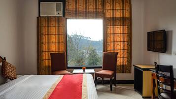 Quarto de hotel com cama, duas cadeiras, uma mesinha, televisão e janela com cortinas.