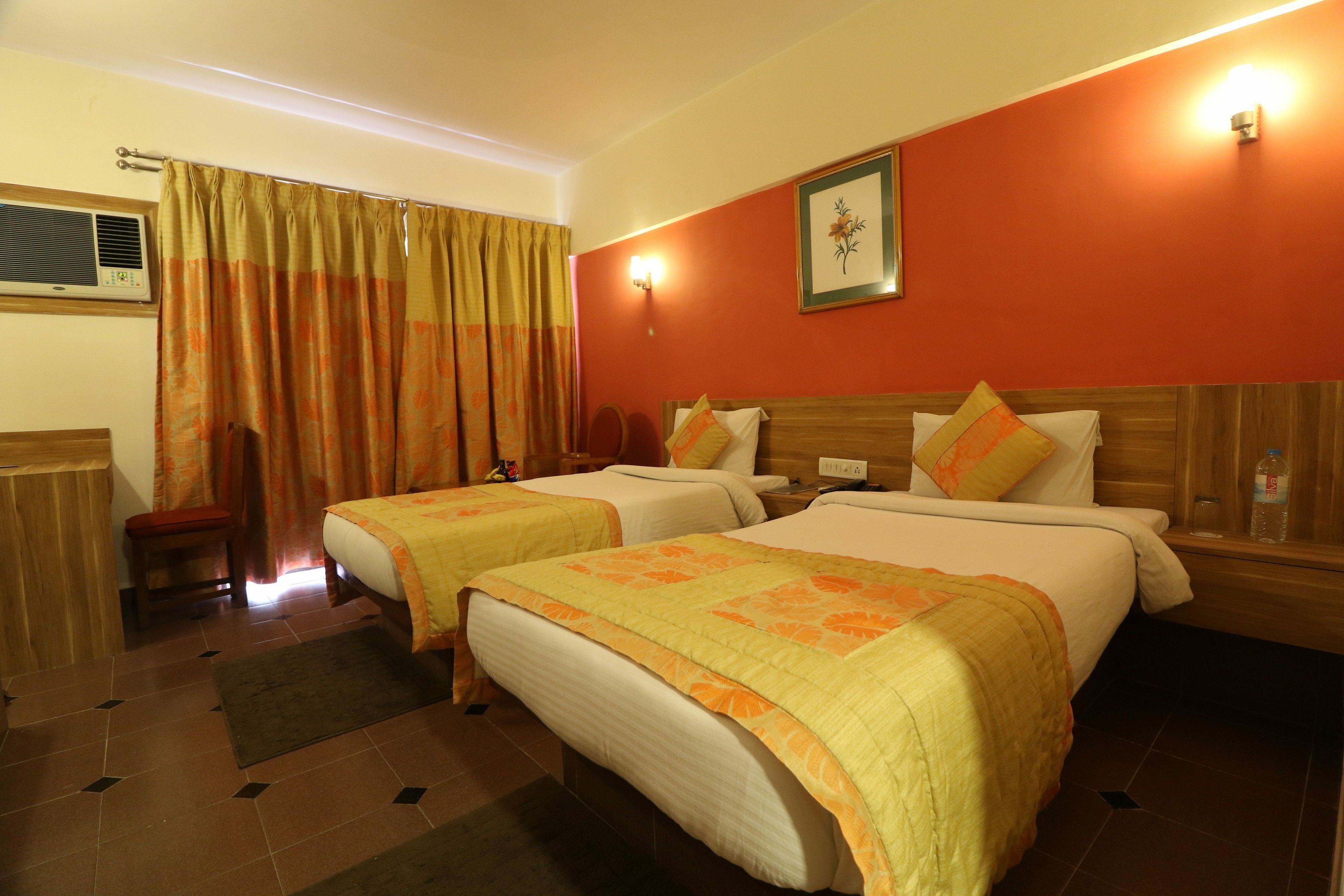 Quarto de hotel com duas camas, cabeceira de madeira, mesa de cabeceira, uma cadeira, janela com cortinas, ar condicionado e luminária fixada na parede.