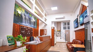 Uma área de recepção com uma mesa de madeira, balcão de atendimento, sala de espera com sofás de couro, televisão na parede e escada para outro andar.