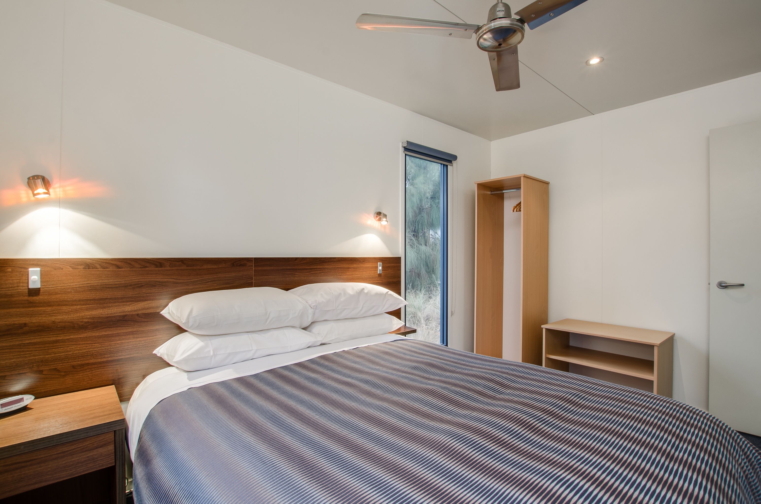 Quarto de hotel moderno com cabeceira de madeira, cama grande com colcha listrada, mesa de cabeceira com termômetro, guarda-roupa e ventilador de teto.