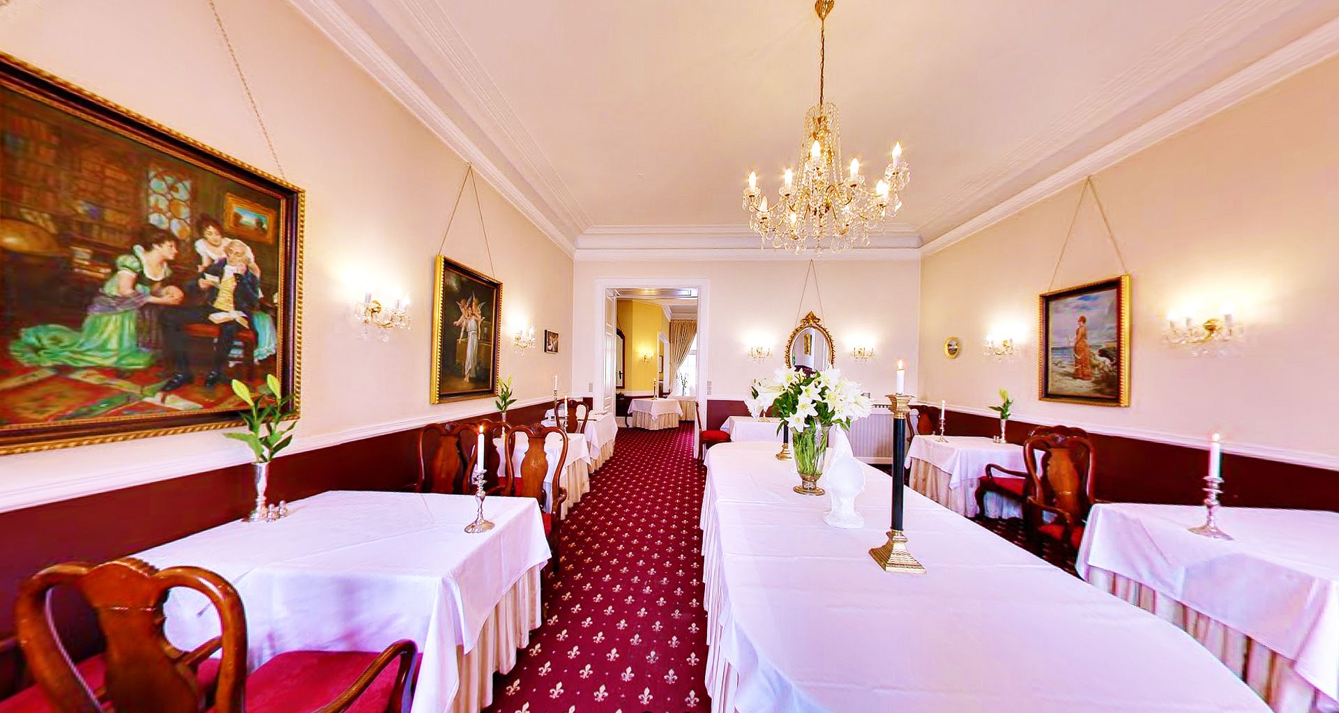 Uma sala de jantar formal com mesas preparadas para o serviço, um candelabro e obras de arte nas paredes.