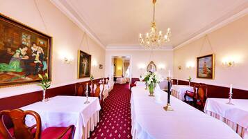 Uma sala de jantar formal com mesas preparadas para o serviço, um candelabro e obras de arte nas paredes.