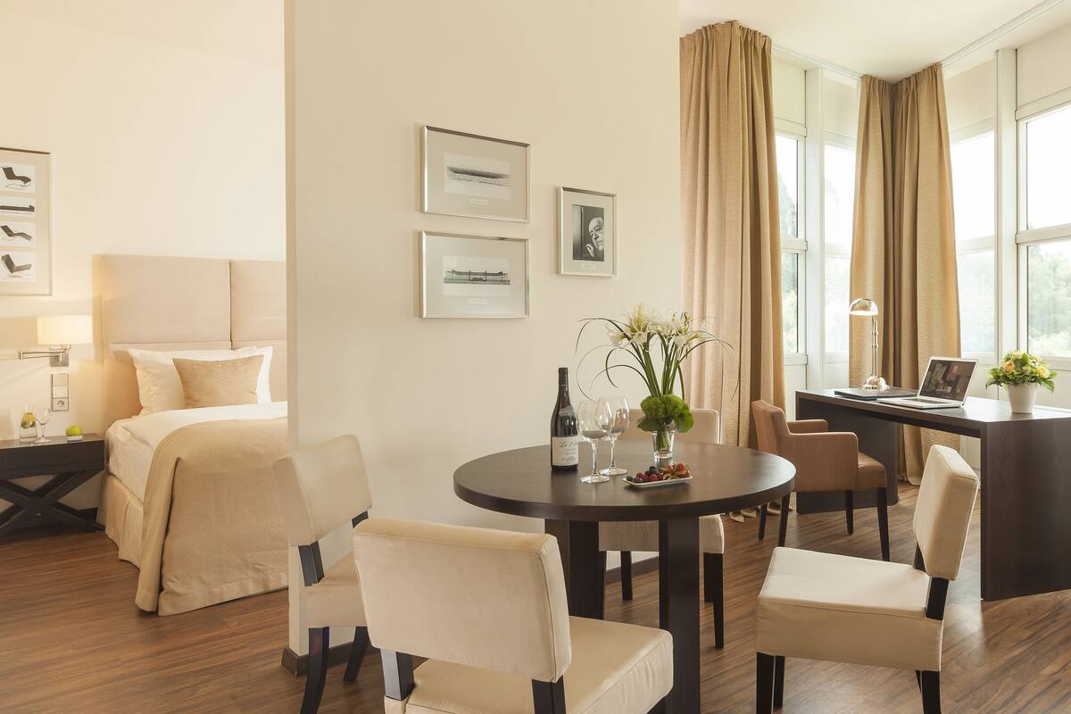 Quarto de hotel com cama, escrivaninha com laptop, mesa de jantar com cadeiras e janela com cortinas.
