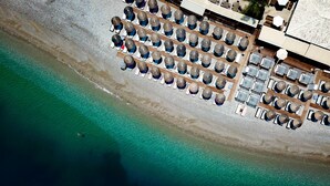 Vista aérea de um hotel à beira-mar, com fileiras de espreguiçadeiras e guarda-sóis, uma área de piscina e um edifício com telhado de azulejos.