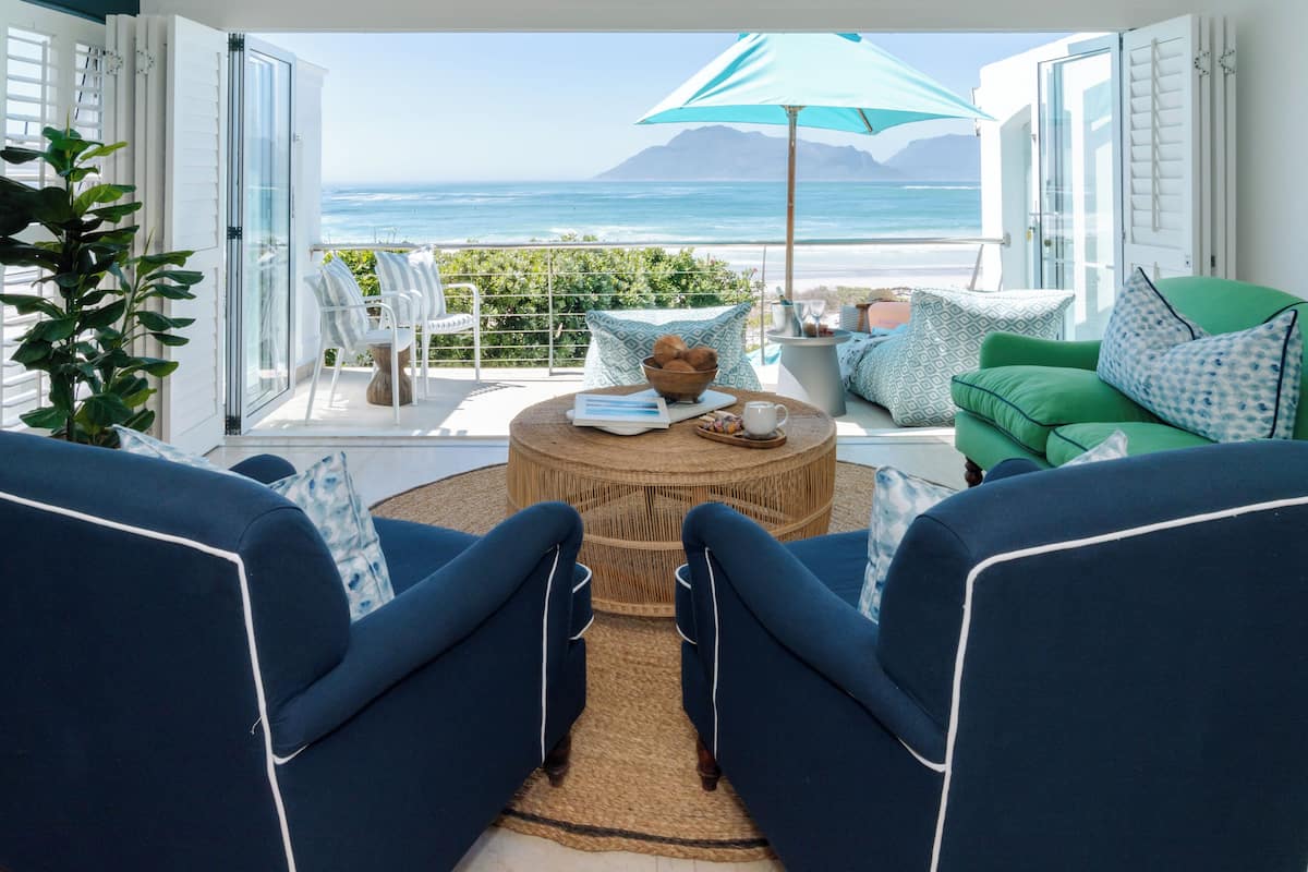 Uma varanda com vista para o oceano, uma mesa de madeira redonda com uma tigela e livros, e duas poltronas azuis com almofadas estampadas.