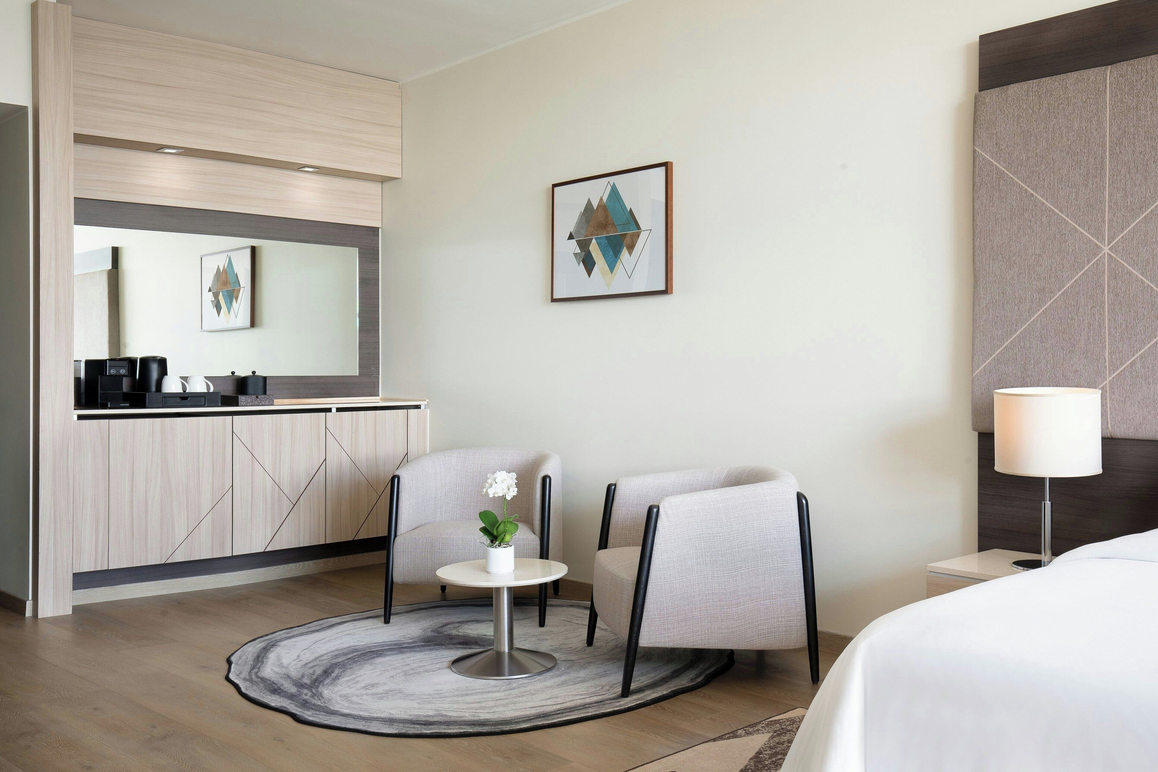 Quarto de hotel moderno com cama, duas poltronas, uma mesa redonda pequena com uma planta, máquina de café, luminária e obras de arte nas paredes.