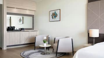 Quarto de hotel moderno com cama, duas poltronas, uma mesa redonda pequena com uma planta, máquina de café, luminária e obras de arte nas paredes.