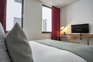 Quarto de hotel com cama, escrivaninha com computador, televisão e janela com cortinas vermelhas.