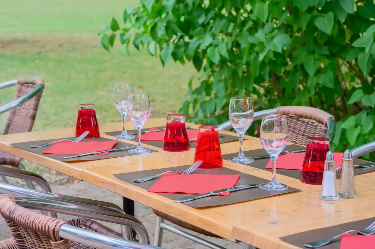 Mesa posta ao ar livre com guardanapos vermelhos, copos de vinho transparentes e saleiro e pimenteiro.