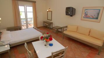 Quarto de hotel com cama, sofá, mesa com um vaso de flores, escrivaninha com abajur, cadeira, televisão e varanda com cadeiras.