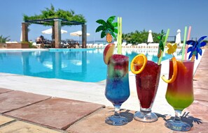 Três cocktails coloridos com guarnições de frutas sobre uma mesa junto à piscina, com uma área de piscina e lounge ao fundo.