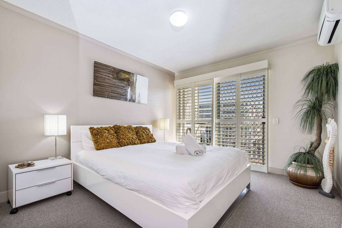 Quarto de hotel moderno com uma cama grande, uma mesa de cabeceira com abajur, um ornamento na parede, uma janela com persianas e um vaso de planta.