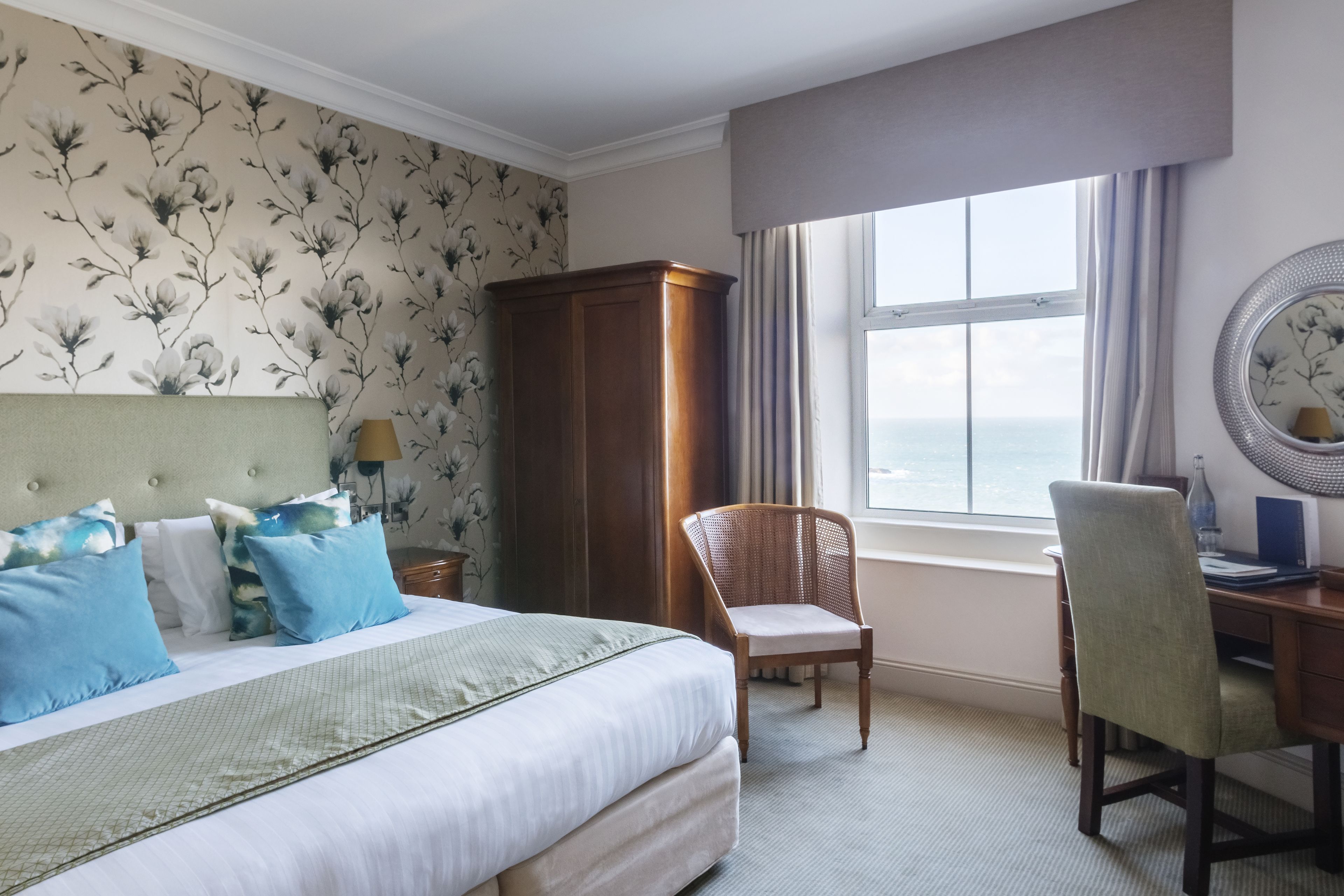 Quarto de hotel com cama, secretária, cadeira, guarda-roupa e vista para o mar através de uma janela.