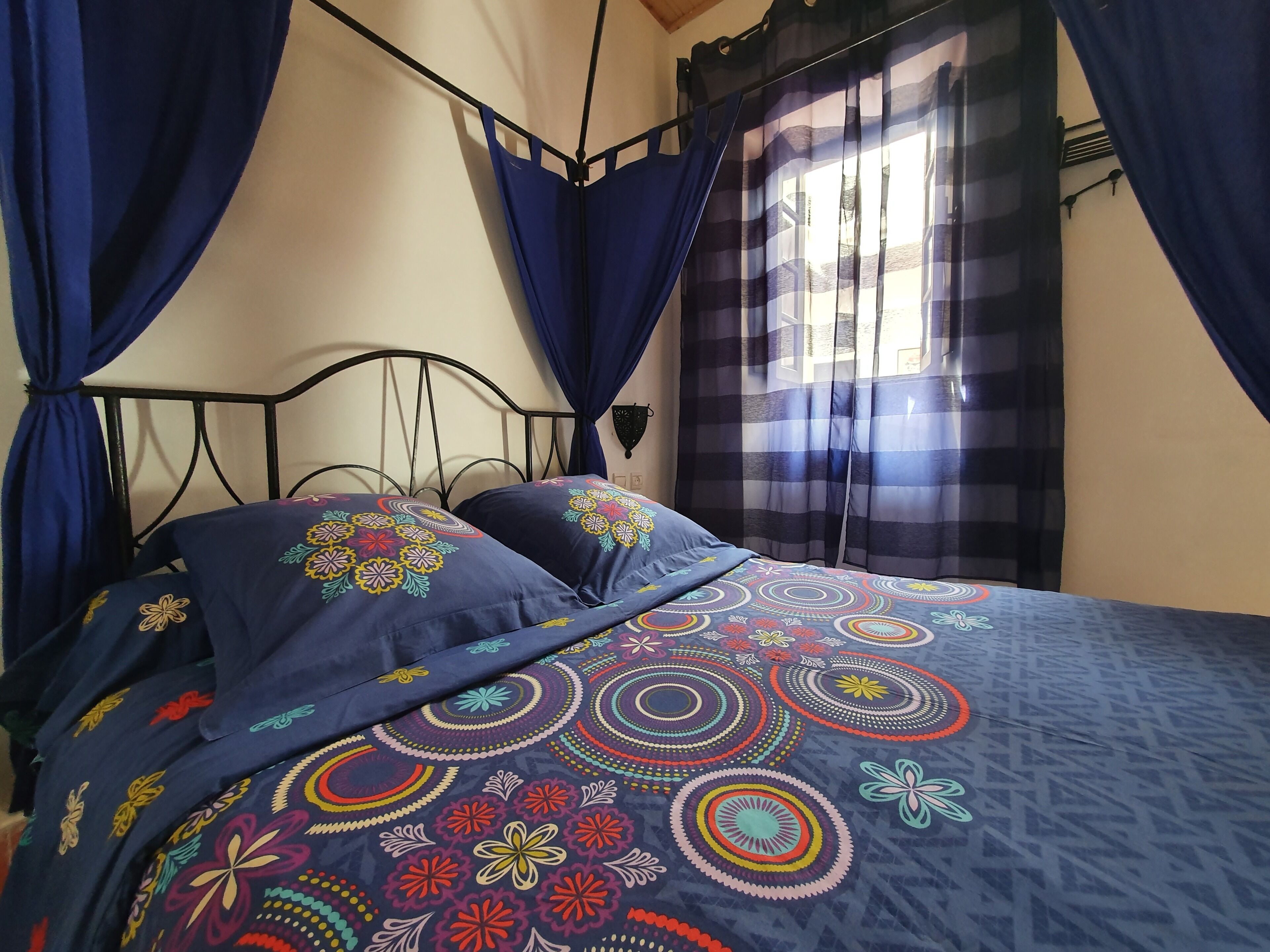 Cama com colcha estampada em azul e cores vibrantes, cabeceira de metal preto e cortinas azuis.