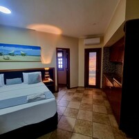Quarto de hotel com cama, pintura de veleiros na parede, área de cozinha e vista através de uma porta aberta.